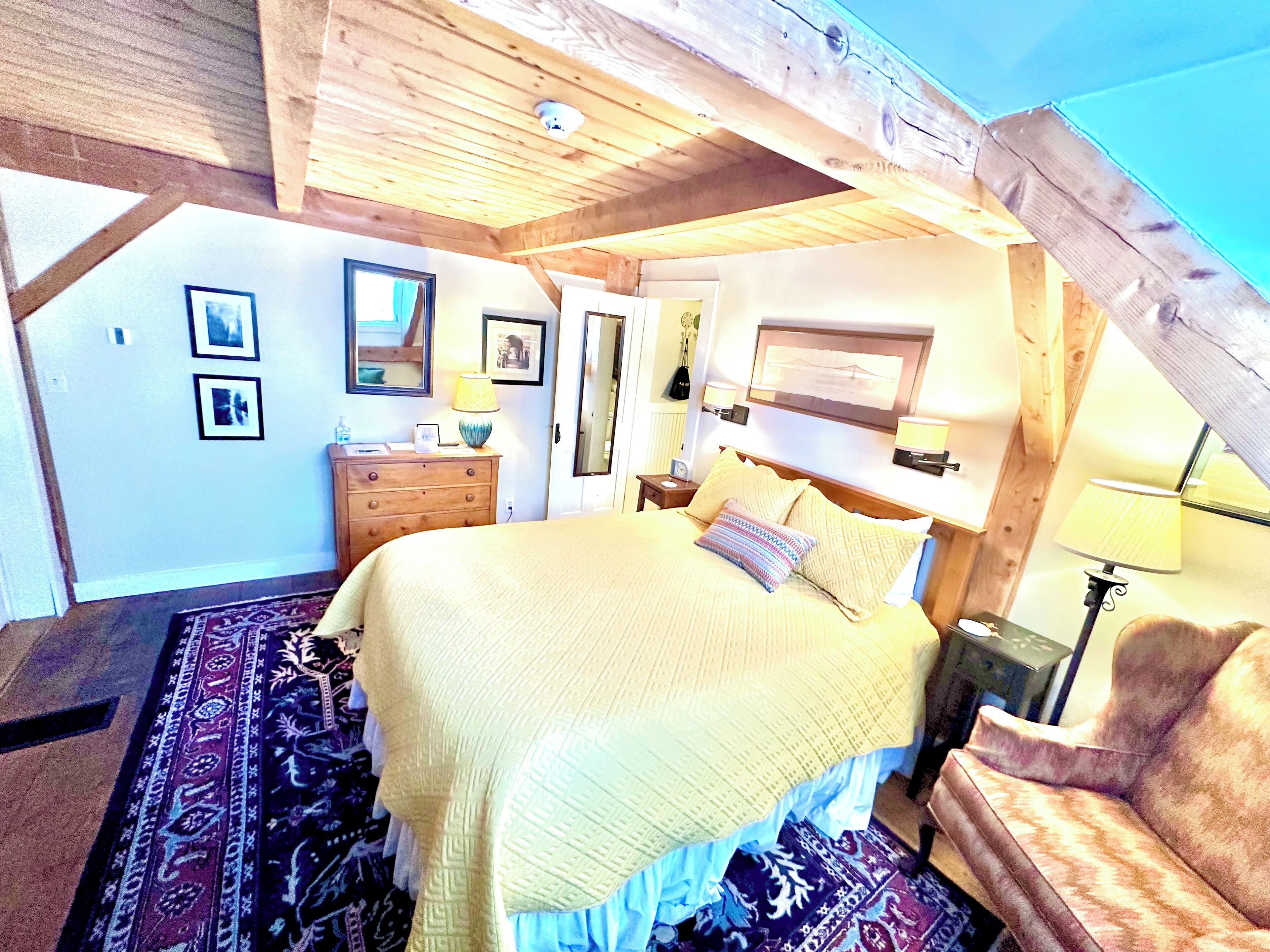 Quarto com cama grande, vigas de madeira no teto, cômoda e mesa de cabeceira com abajur.