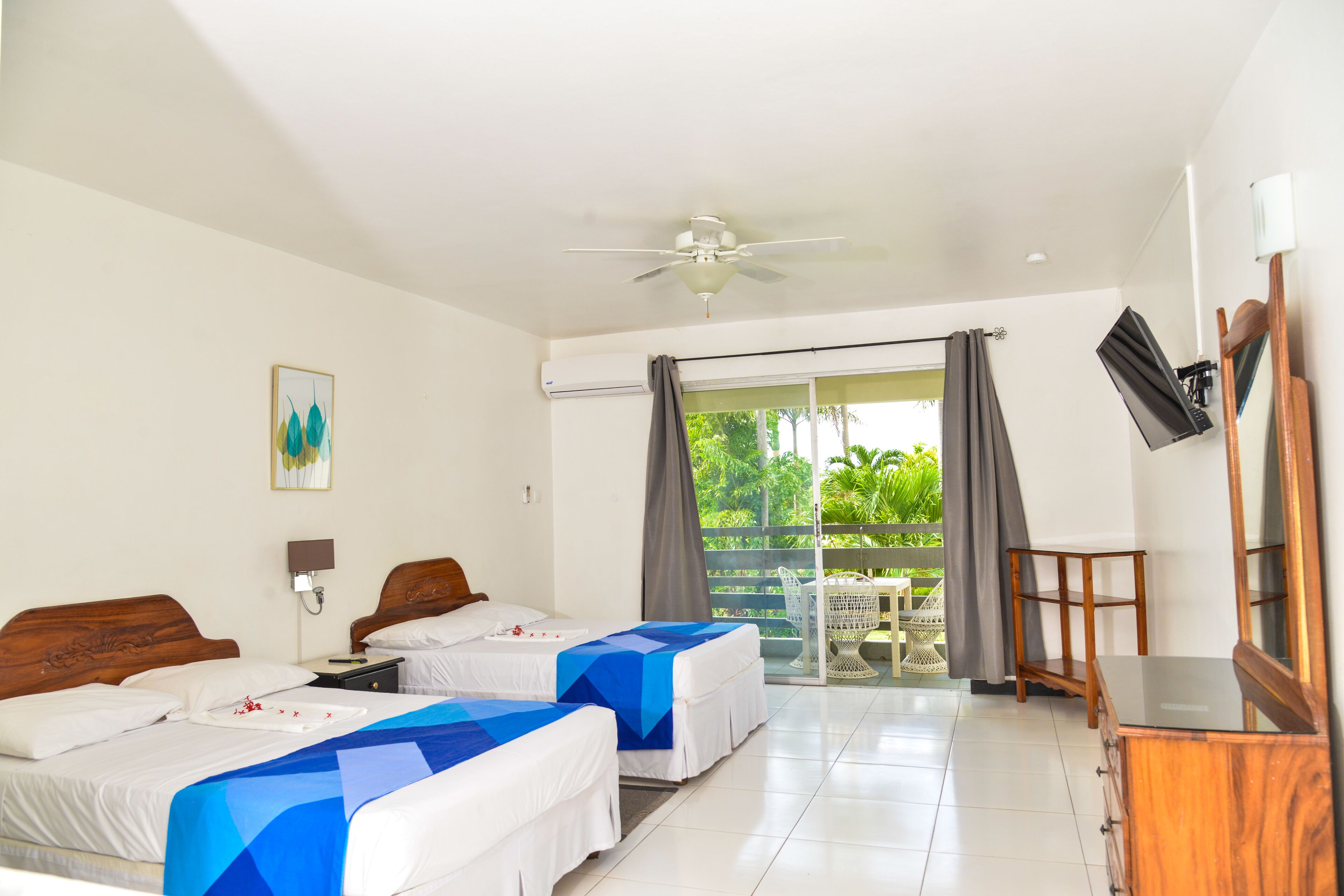 Quarto de hotel com duas camas, ventoinha no teto, televisão e varanda com vista para o verde.