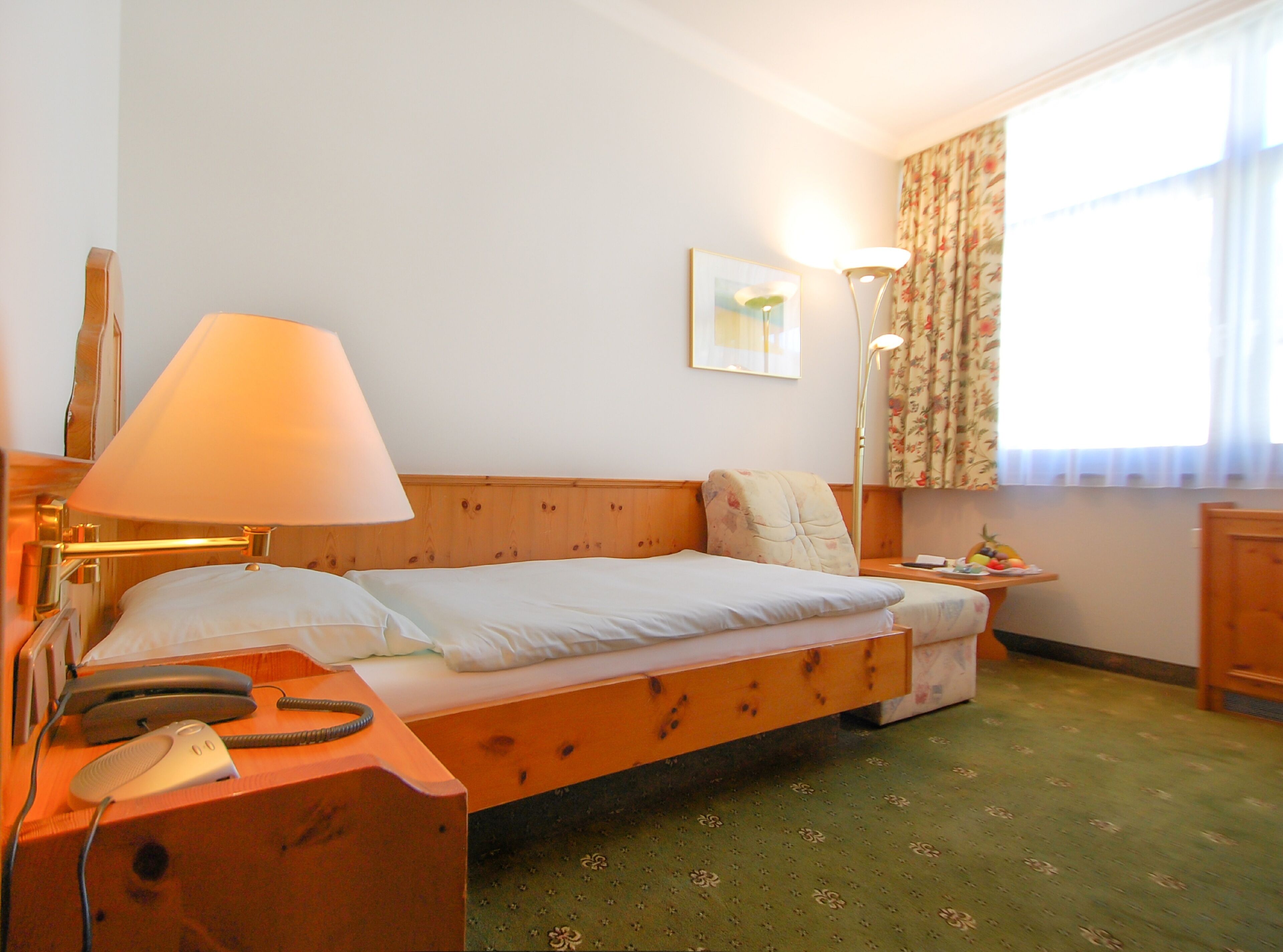 Quarto de hotel com uma cama de madeira, mesa de cabeceira com abajur, telefone e pequena mesa com frutas.