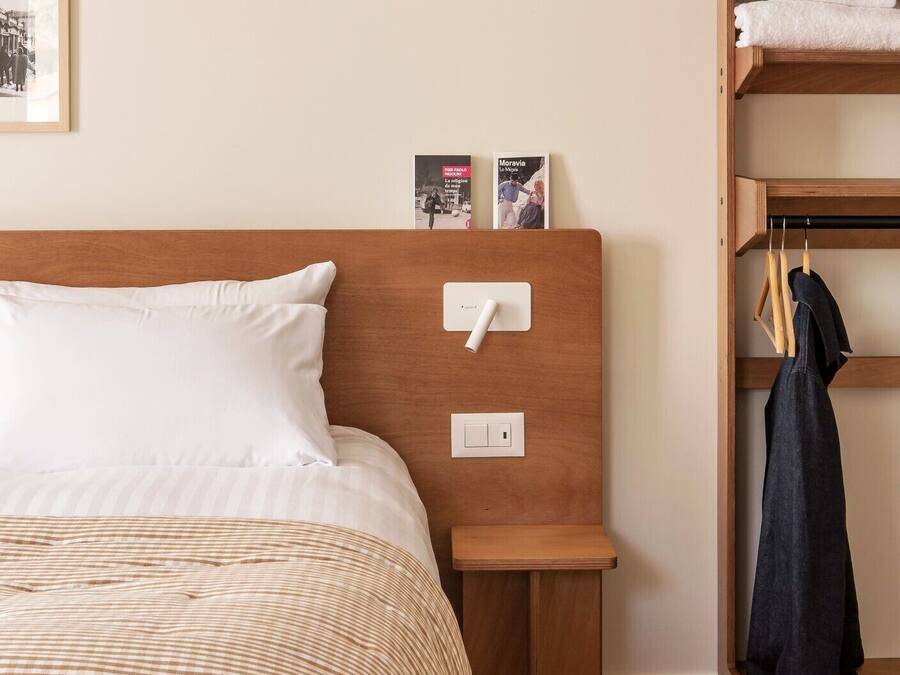 Uma cama bem arrumada com manta xadrez, mesa de cabeceira com interruptor de luz, pequena estante com livros e guarda-roupa de madeira com roupas penduradas.