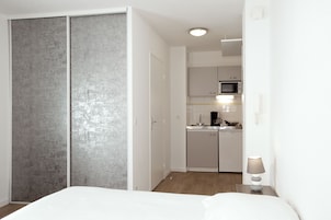 Quarto com cama, guarda-roupa e uma kitchenette ao fundo.