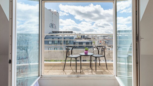 Varanda com mesa, cadeiras, planta em vaso e vista de prédios e nuvens.