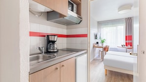 Cozinha moderna com fogão embutido, máquina de café e pia. Há uma cama com cabeceira branca e mesa de cabeceira com abajur.