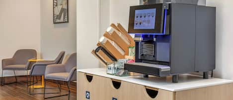 Máquina de café moderna com visor digital, moedor de café e dispensador de água. Dois cadeiras cinzentas e uma pequena mesa em primeiro plano.