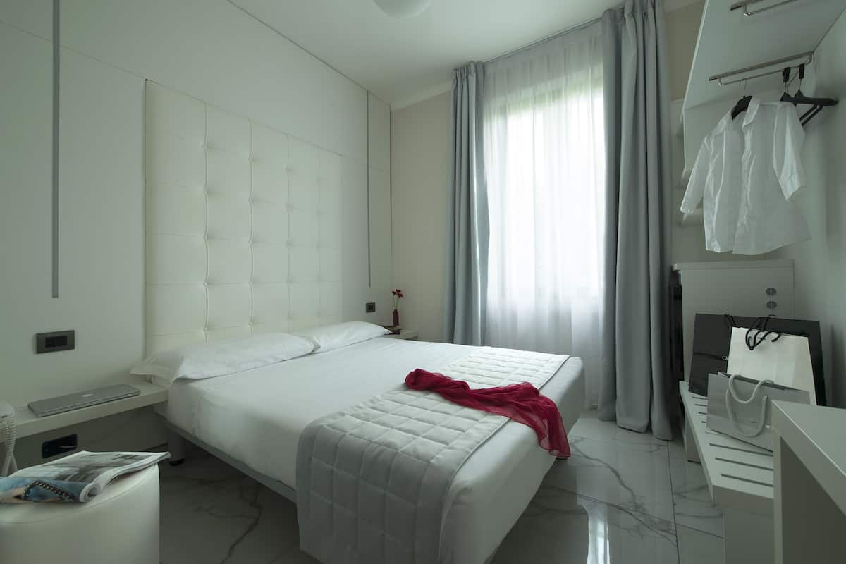 Quarto de hotel moderno com uma cama grande, mesa de cabeceira com abajur, pequena escrivaninha com cadeira e janela com cortinas.