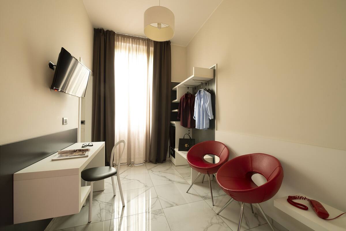 Quarto de hotel moderno com uma escrivaninha, duas cadeiras vermelhas, uma TV fixada na parede, um cabideiro com roupas e uma janela grande com cortinas.