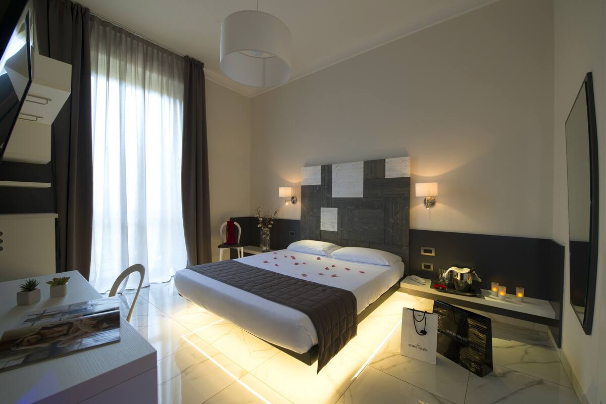 Quarto de hotel moderno com uma cama bem arrumada, uma mesa de cabeceira com abajur, uma escrivaninha com cadeira e um espelho grande na parede.