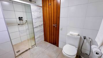 Uma casa de banho com cabine de chuveiro em vidro, porta de madeira, sanita e um cesto de lixo.