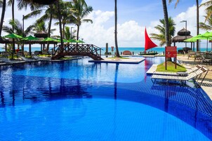 Piscina com espreguiçadeiras e guarda-sóis, rodeada por palmeiras e vista para o oceano.