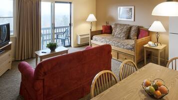 Quarto de hotel acolhedor com um sofá vermelho, uma mesa de jantar de madeira com uma cesta de frutas, uma televisão pequena e uma varanda com vista.