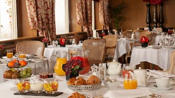 Mesa posta para o café da manhã, com diversas opções de comida e bebida, incluindo frutas, doces e refrescos, sobre toalhas brancas e cadeiras de vime.
