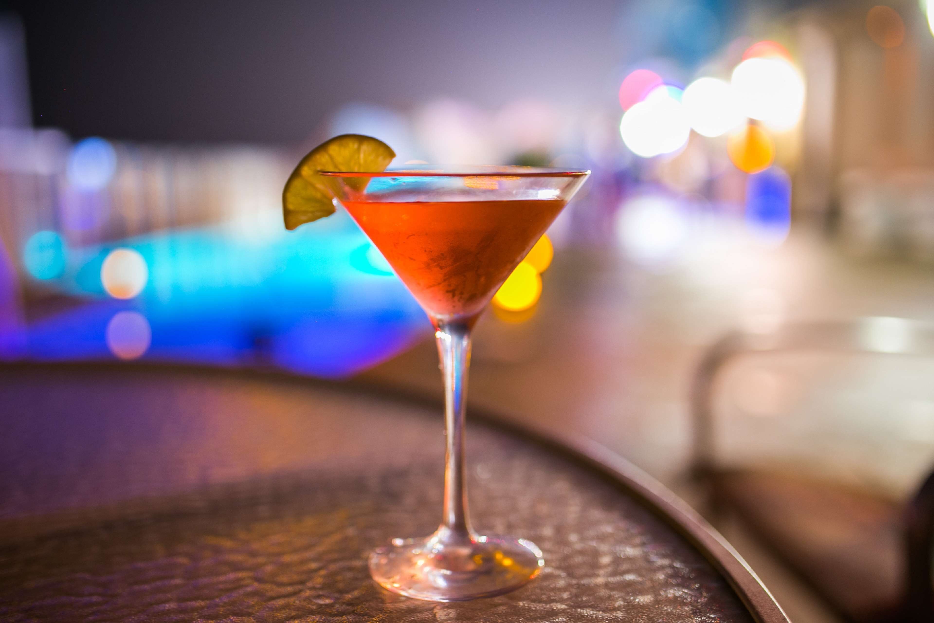 Um cocktail num copo de martini com um toque de limão sobre uma mesa, com luzes desfocadas ao fundo.