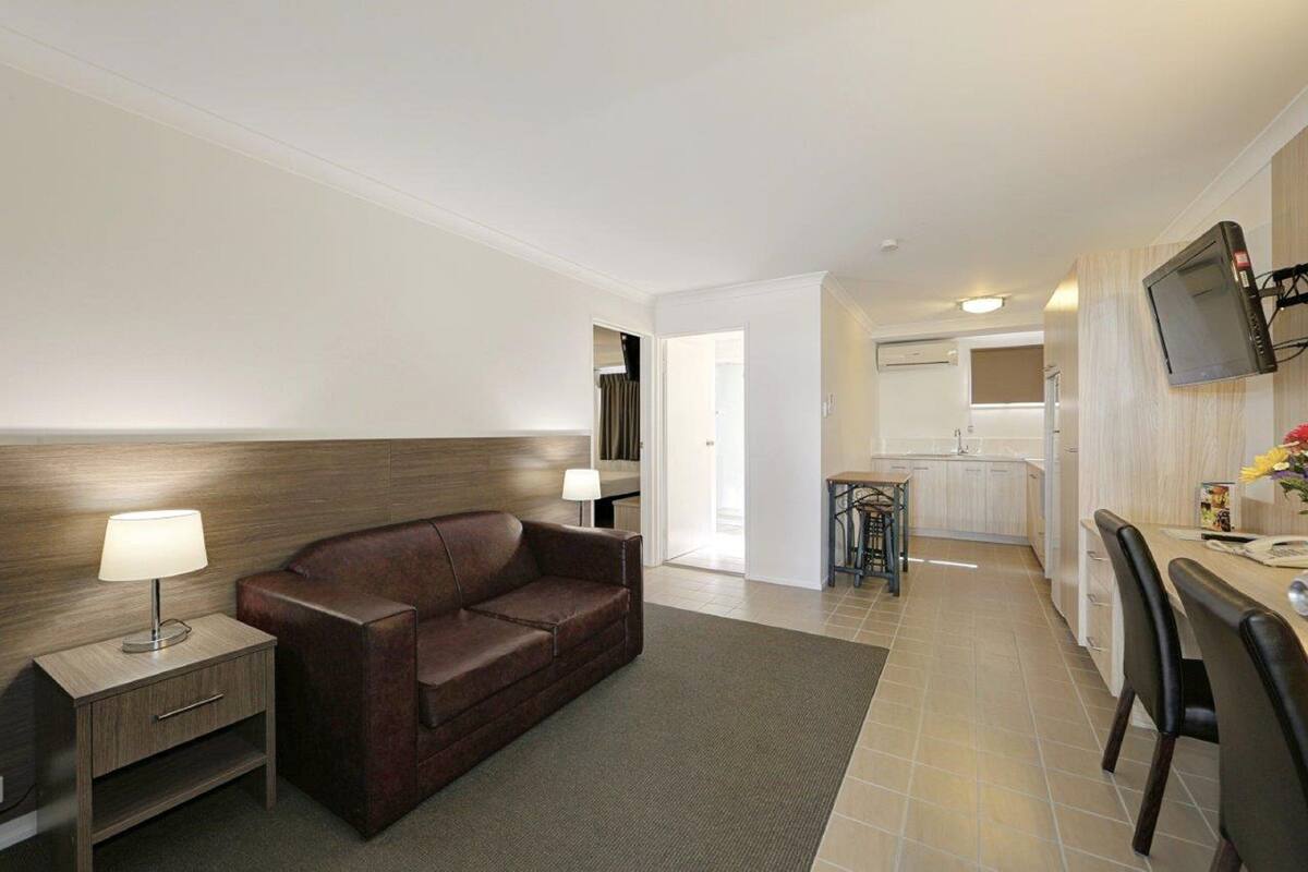 Quarto de hotel moderno com sofá de couro marrom, mesa de cabeceira com luminária, cozinha compacta e televisão fixada na parede.