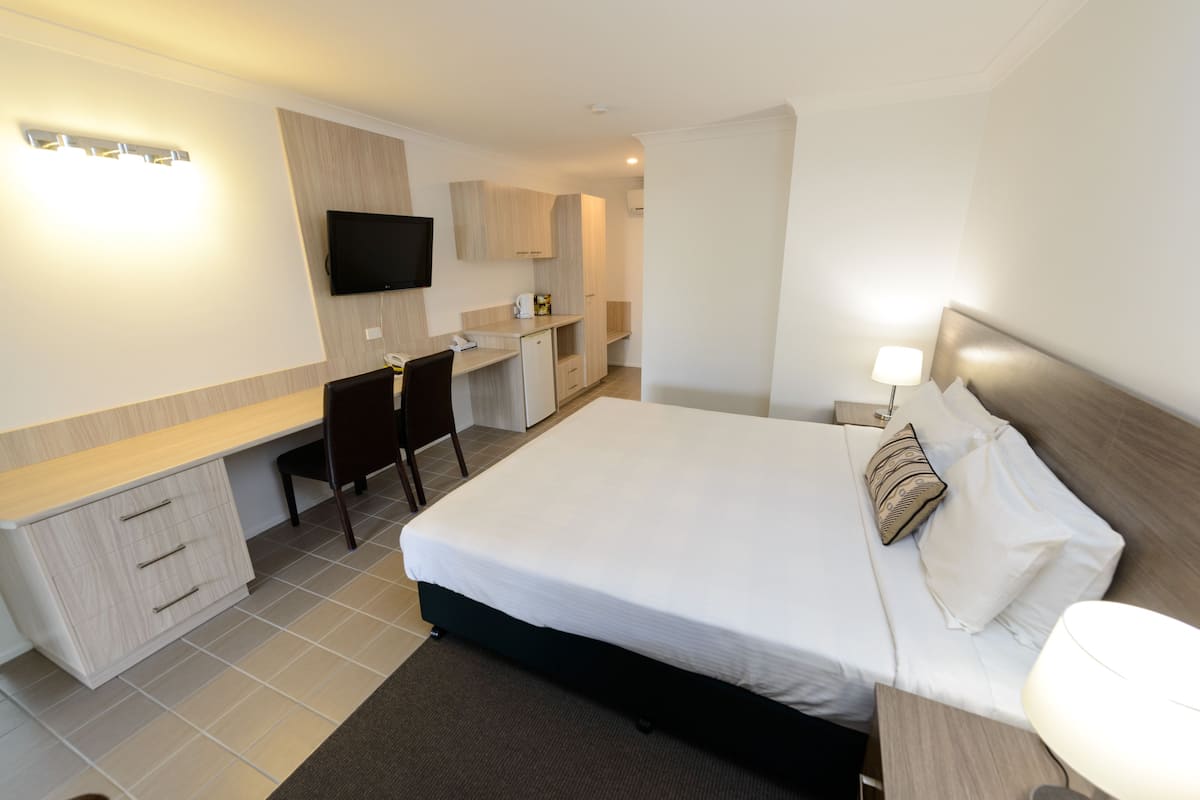Quarto de hotel com cama, escrivaninha, cadeira e televisão fixada na parede.