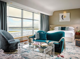 Quarto de hotel com uma grande janela, uma cama, um sofá verde-água, uma mesa de centro de vidro e vista para a praia.