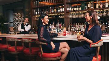Uma área de bar com duas mulheres sentadas ao balcão, um bartender atrás do balcão e prateleiras repletas de garrafas e copos.