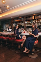 Uma área de bar com duas mulheres sentadas ao balcão, um bartender atrás do balcão e prateleiras repletas de garrafas e copos.