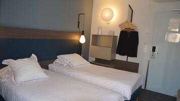 Quarto de hotel com duas camas, uma mesinha de cabeceira, prateleira na parede, cabideiro e luminária.