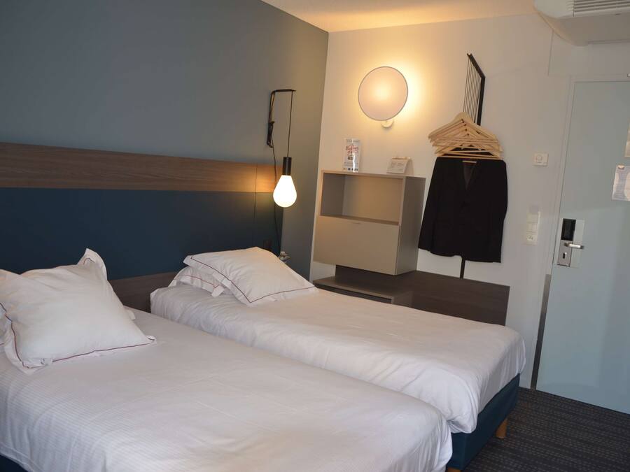 Quarto de hotel com duas camas, uma mesinha de cabeceira, prateleira na parede, cabideiro e luminária.
