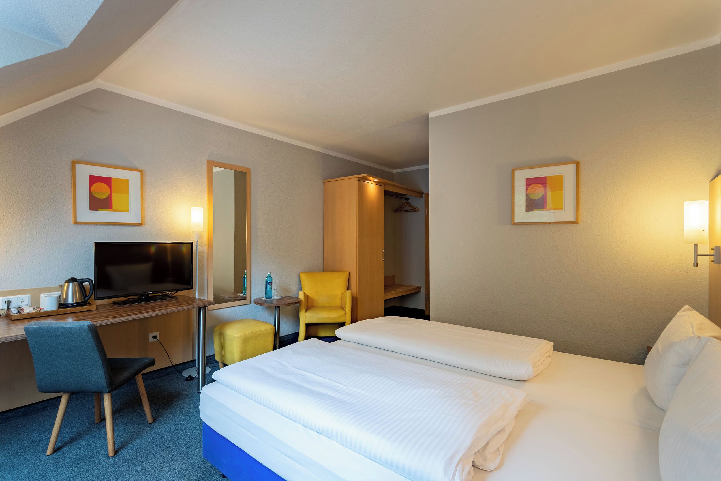 Quarto de hotel com cama, escrivaninha com televisão, cadeira e guarda-roupa.