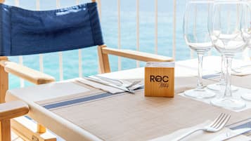 Mesa de jantar com um jogo americano, uma caixa de madeira com a inscrição "ROC" e duas taças de vinho numa varanda com vista para o mar.