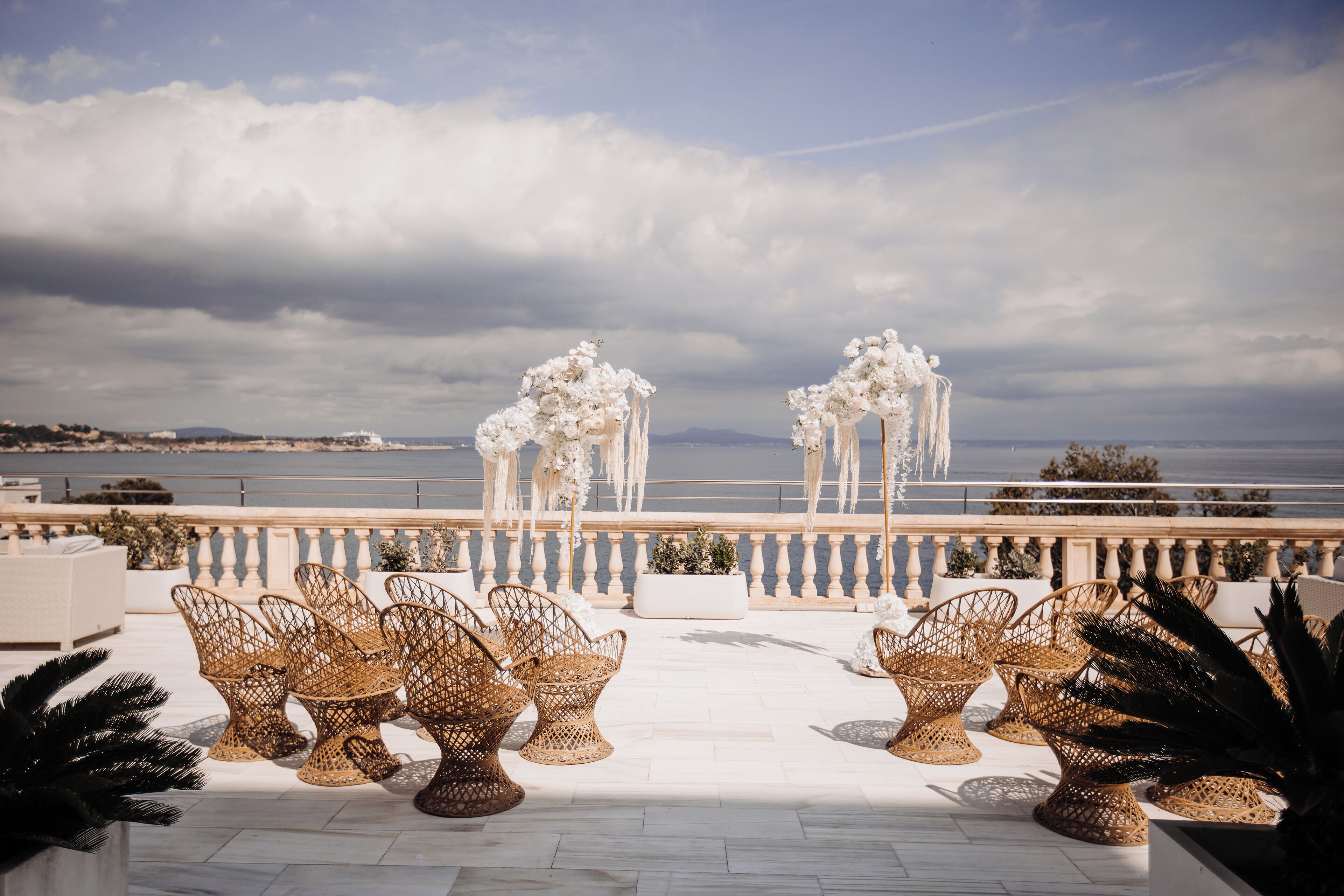 Terraço com cadeiras de vime e decorações florais brancas, com vista para um corpo de água.