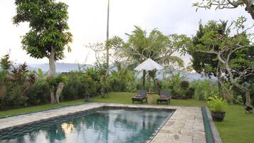 Piscina retangular com borda de pedra, rodeada por vegetação exuberante e duas espreguiçadeiras com guarda-sóis.