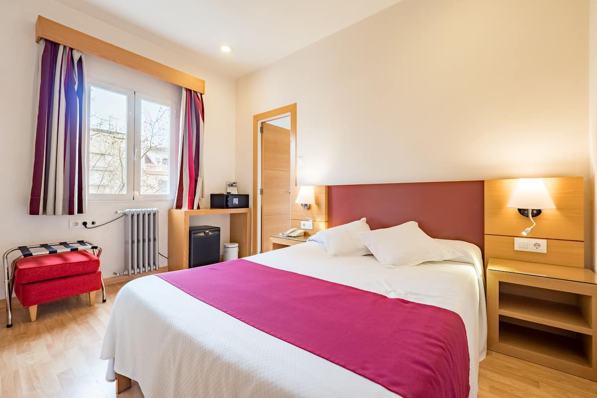 Quarto de hotel com uma cama grande, um banco vermelho, cabeceira de madeira, mesas de cabeceira com abajures e uma janela com cortinas.