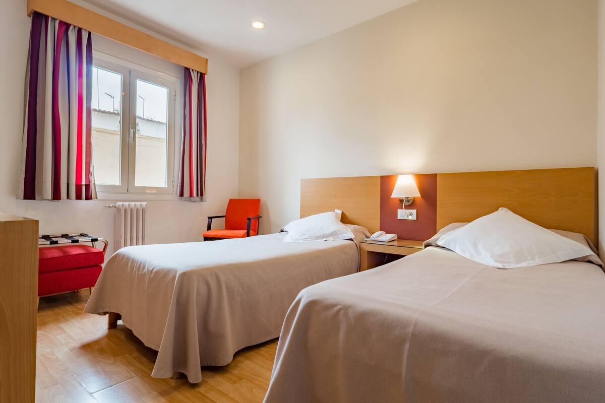 Quarto de hotel com duas camas, uma cadeira vermelha, cabeceira de madeira e janela com cortinas.
