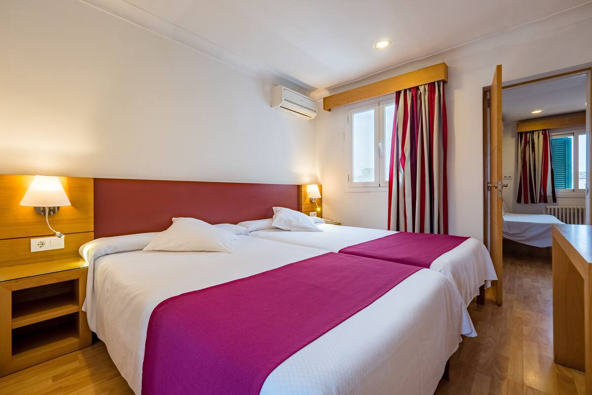 Quarto de hotel com duas camas, uma cabeceira de madeira, uma mesa de cabeceira com abajur e uma janela com cortinas listradas.