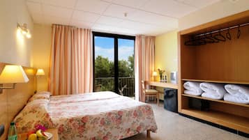 Quarto de hotel com uma cama, uma escrivaninha, uma cadeira e um guarda-roupa com roupas penduradas.