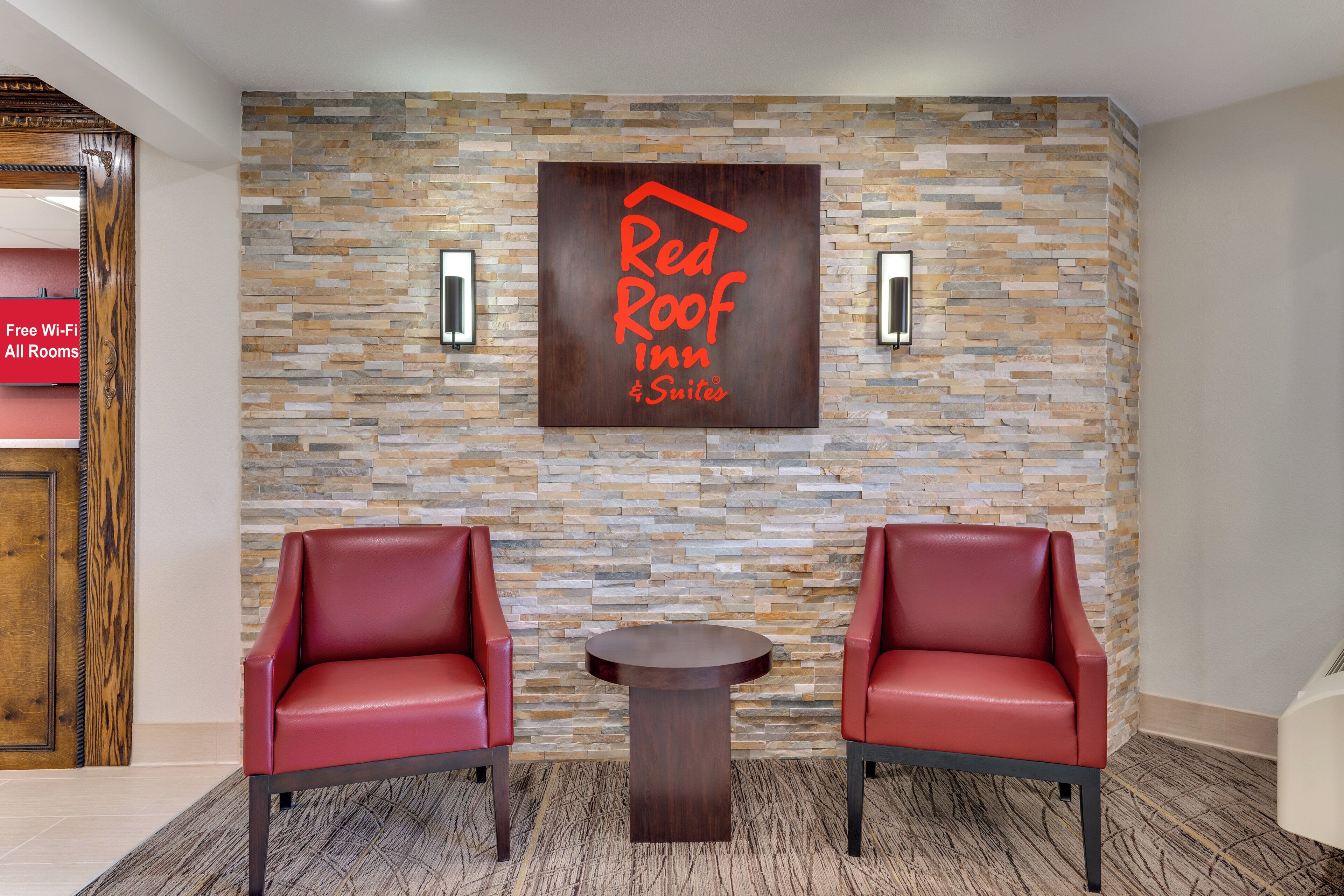 Um hall com parede de pedra, duas poltronas de couro vermelho, uma mesa pequena e uma placa com a inscrição 'Red Roof Inn Suites'.