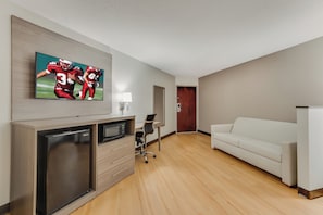 Quarto de hotel com televisão de tela plana na parede, micro-ondas embutido, armário de madeira, sofá branco e piso de madeira.