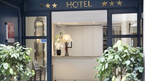 Entrada de hotel com balcão de recepção, placa com o nome do hotel e duas plantas em vasos.