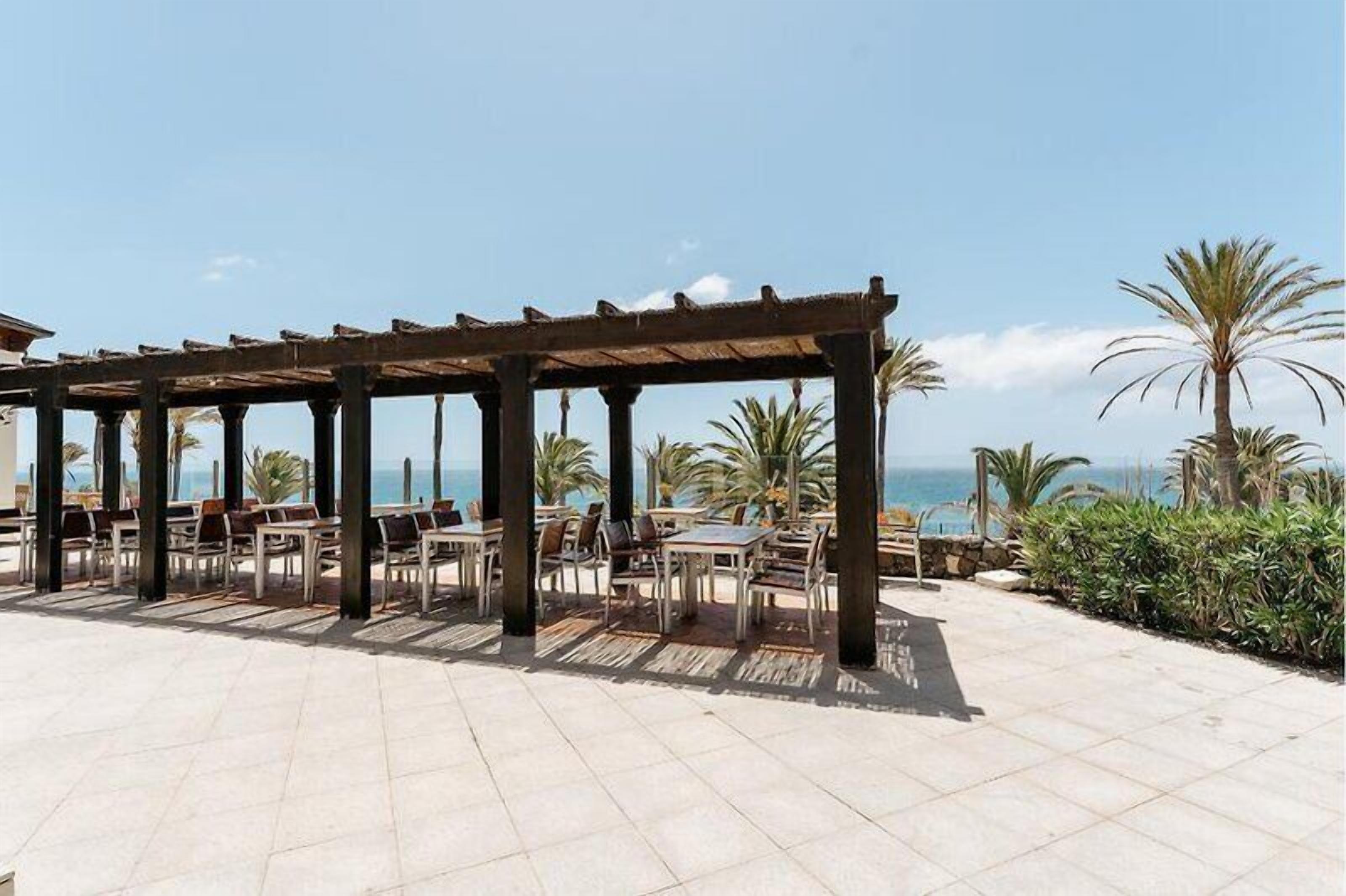 Área externa com mesas e cadeiras, estrutura de pérgola e vista para o mar.