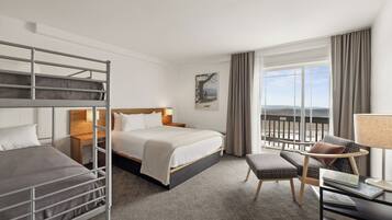 Quarto de hotel com beliche, uma cama de solteiro, escrivaninha com cadeira, vista para o exterior e janela com cortinas.