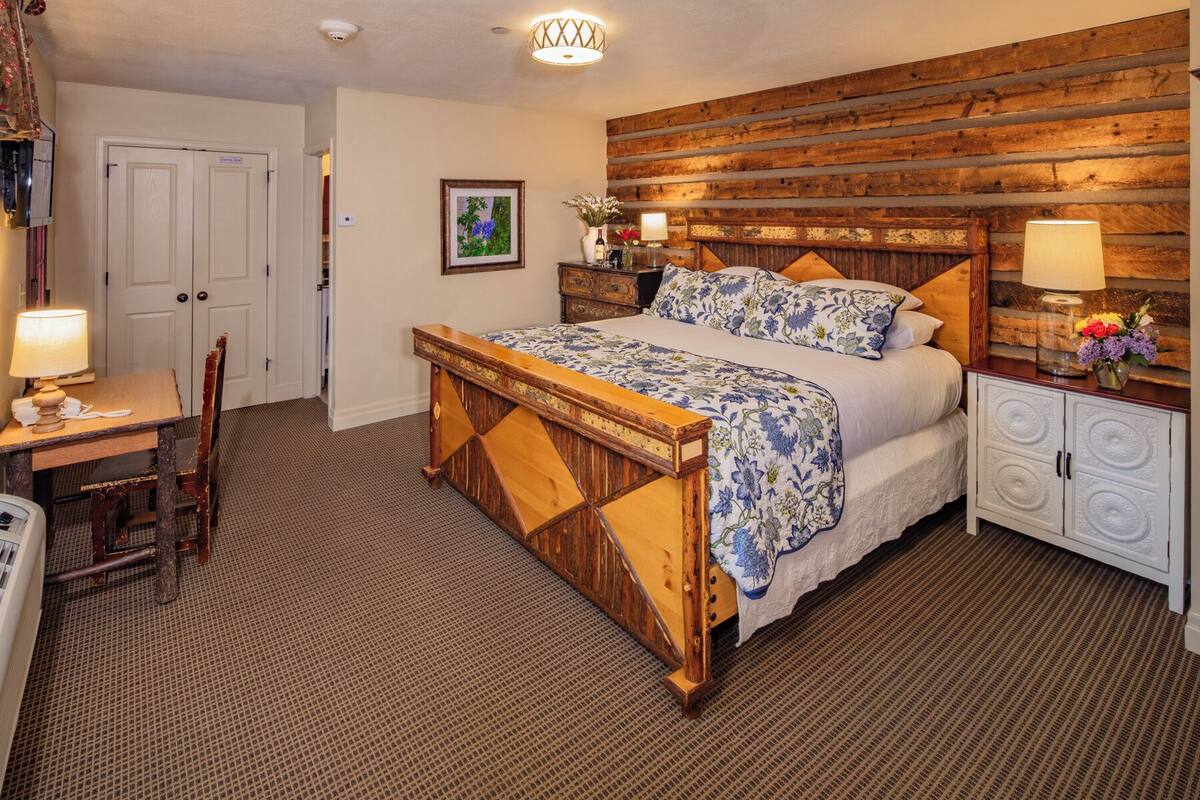 Quarto com cabeceira de madeira, cama com roupa de cama floral, mesa de cabeceira com abajur, cômoda e uma cadeira.