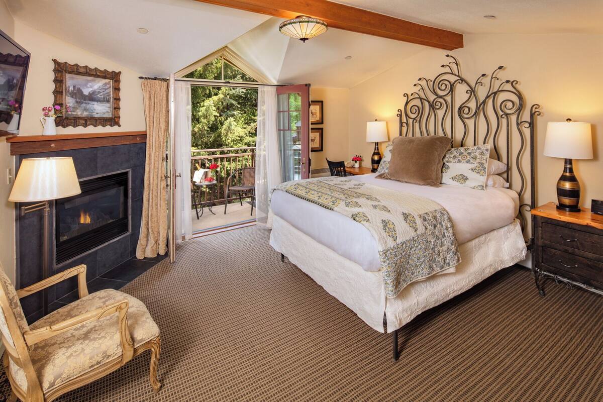 Quarto com cama grande, lareira, varanda com mesa e cadeiras, e vista para o verde.