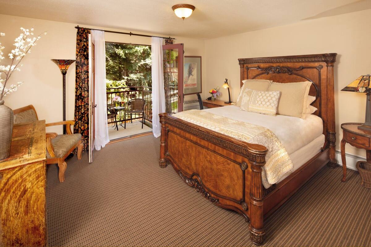 Um quarto tradicional com uma cama de madeira com dossel, um espaço de estar com uma poltrona e uma mesinha, e uma varanda com vista para o verde.