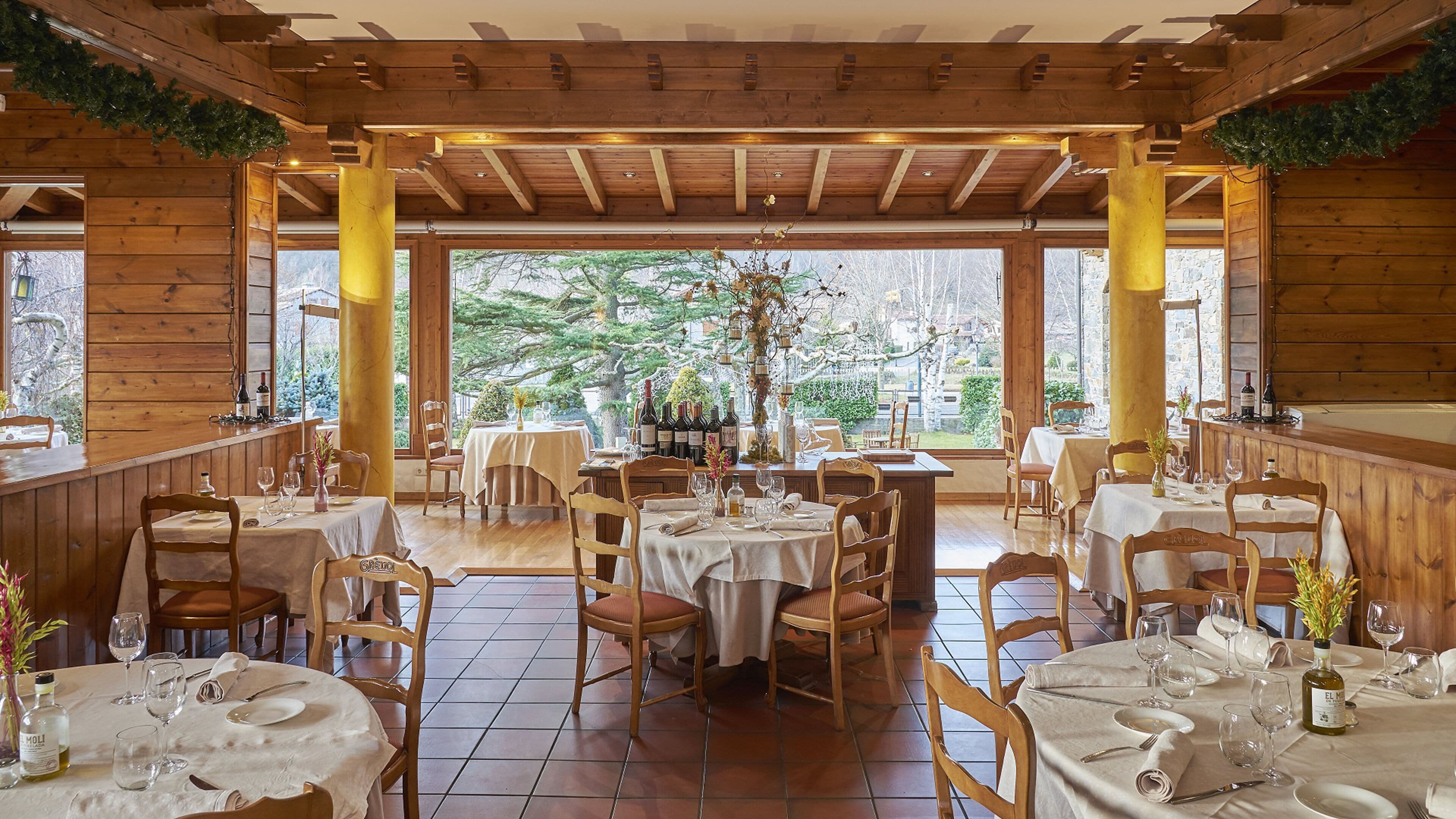 Restaurante com interiores em madeira, várias mesas e cadeiras, e grandes janelas com vista para árvores e uma parede de pedra.