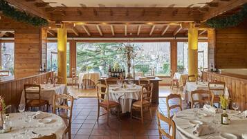 Restaurante com interiores em madeira, várias mesas e cadeiras, e grandes janelas com vista para árvores e uma parede de pedra.