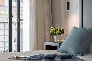 Quarto com cama, mesa de cabeceira com um vaso de flores, janela com cortinas e vista para uma varanda.