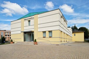 Um edifício com uma placa que diz 'HOTEL SAN CARLO'. Possui um telhado plano, uma entrada grande com portas de vidro e um pátio pavimentado.