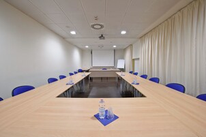 Uma sala de conferências com uma mesa comprida, cadeiras azuis, um quadro branco e uma tela de projetor.