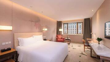 Quarto de hotel com uma cama grande, uma escrivaninha, uma cadeira, uma mesinha e uma janela com vista para edifícios.