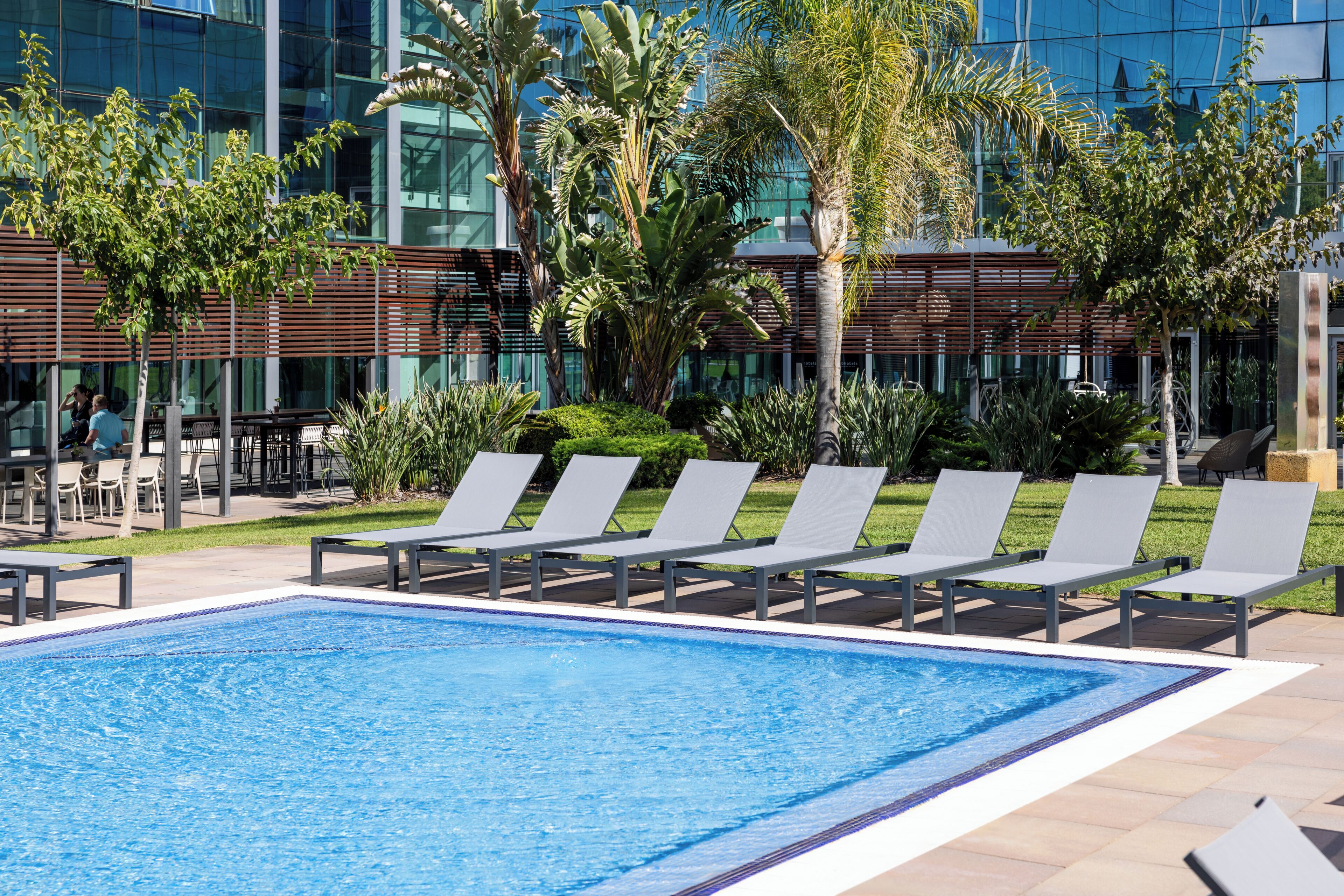 Piscina com cadeiras de descanso, cercada por palmeiras e um edifício moderno.