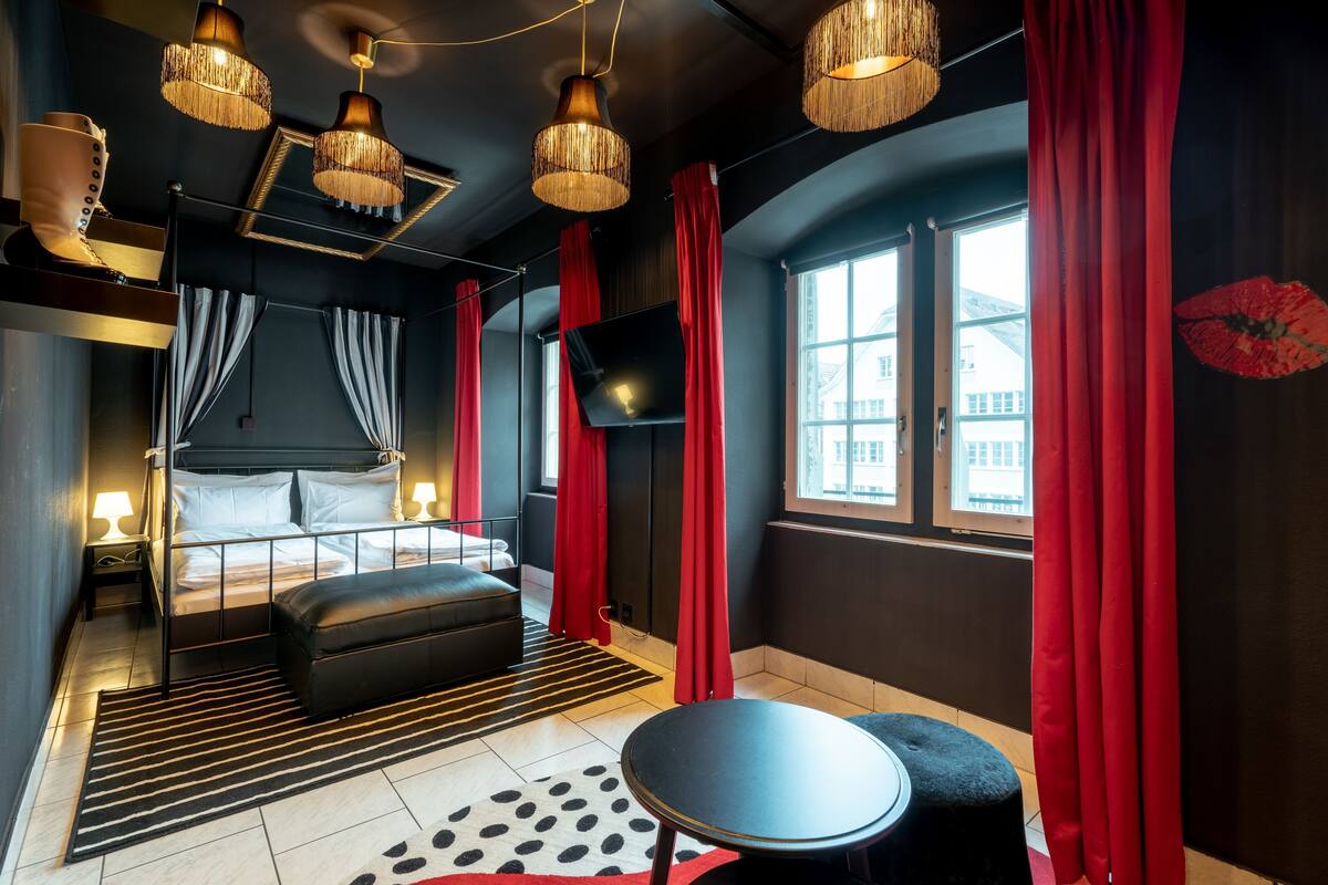 Quarto de hotel moderno com uma cama, uma TV de tela plana, uma mesa redonda e cortinas vermelhas.