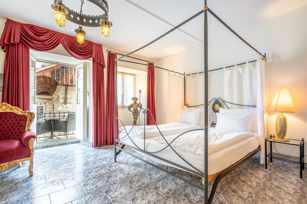 Uma cama de dossel com lençóis brancos, uma cadeira estofada vermelha, uma mesinha com abajur e vista para um pátio externo com mesa e cadeiras.
