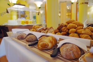 Mesa de buffet com diversos pães e produtos de pastelaria, incluindo croissants e pão, dispostos em tábuas de madeira.