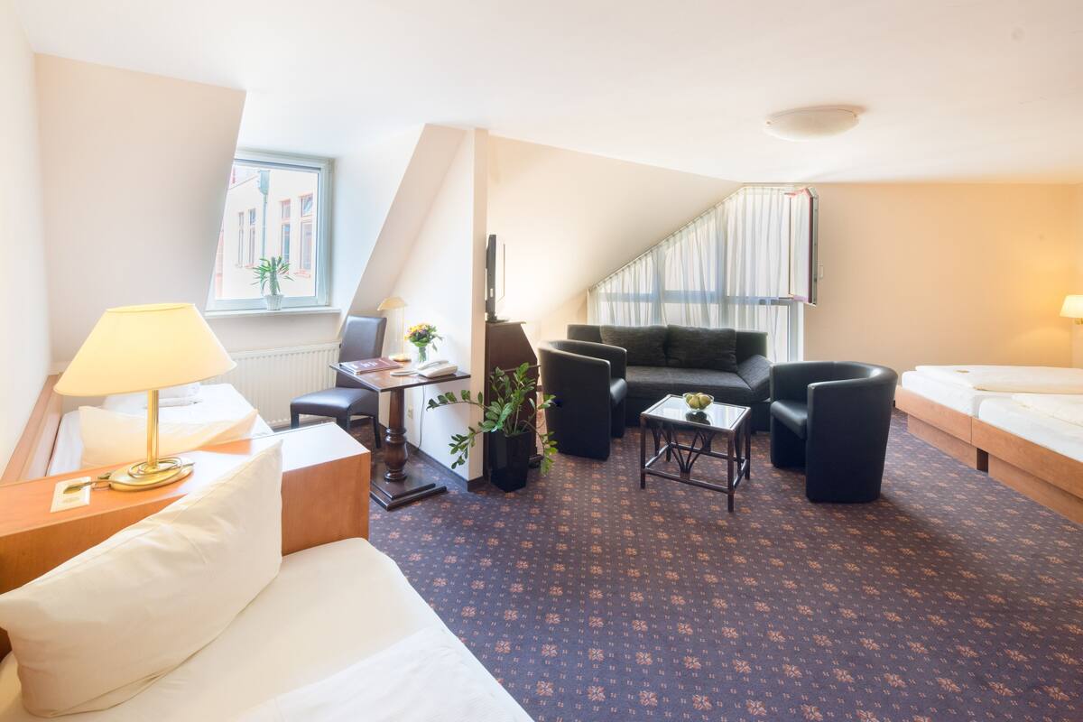 Quarto de hotel com sofá, poltronas, uma mesinha, abajur, planta em vaso e vista da cidade pela janela.